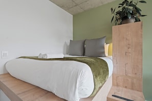 Quarto com cama, roupa de cama verde e branca, cabeceira de madeira, planta em vaso e almofada listrada.