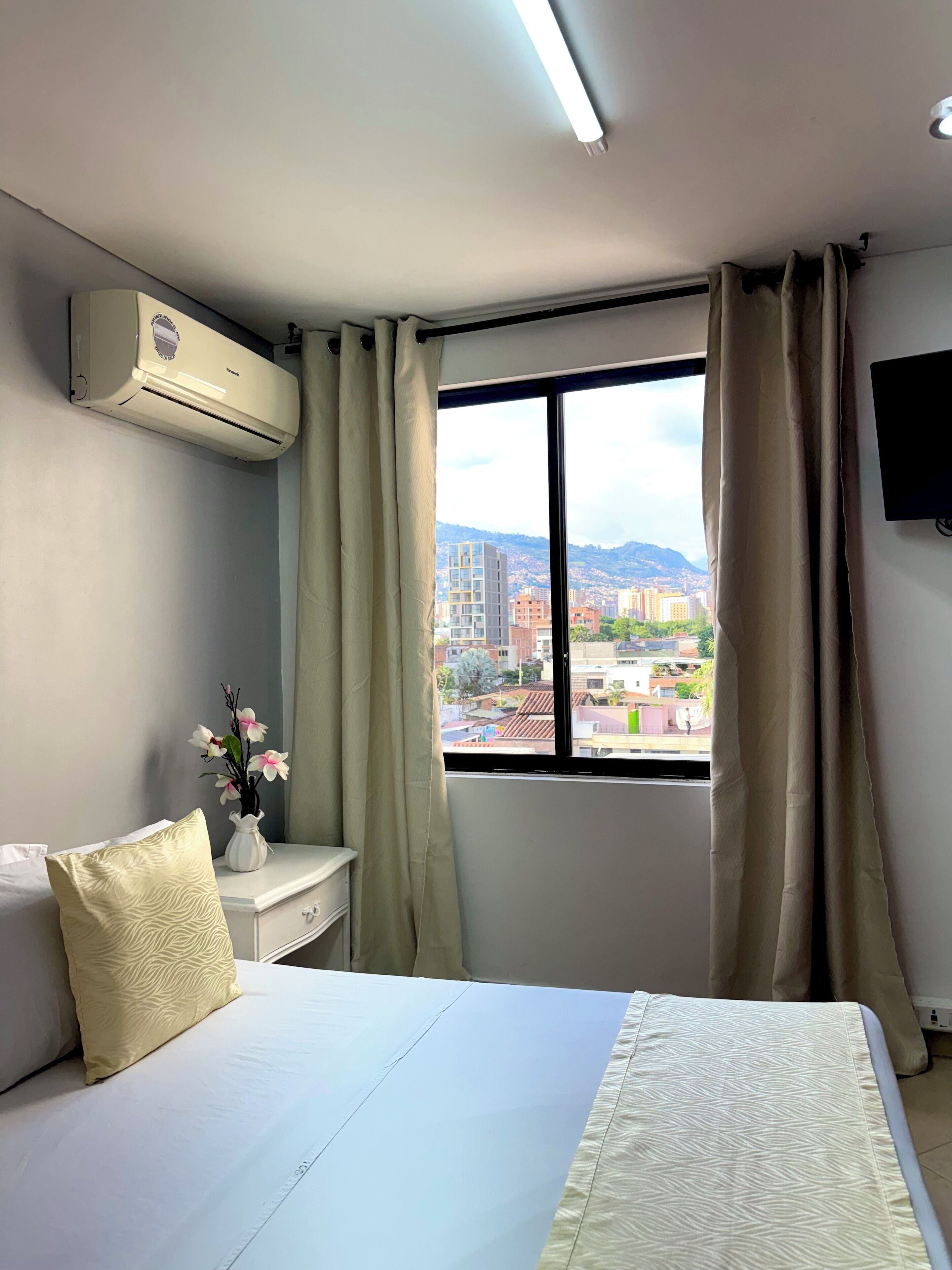 Quarto de hotel com uma cama grande, mesa de cabeceira com um vaso de flores, ar condicionado, janela com cortinas e vista para a cidade.