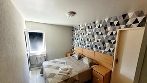 Quarto com cama, mesa de cabeceira, cômoda, janela e parede com padrão geométrico.