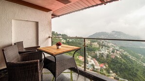 Varanda com mobiliário de vime e mesa de madeira, com vista para uma paisagem montanhosa com edifícios.