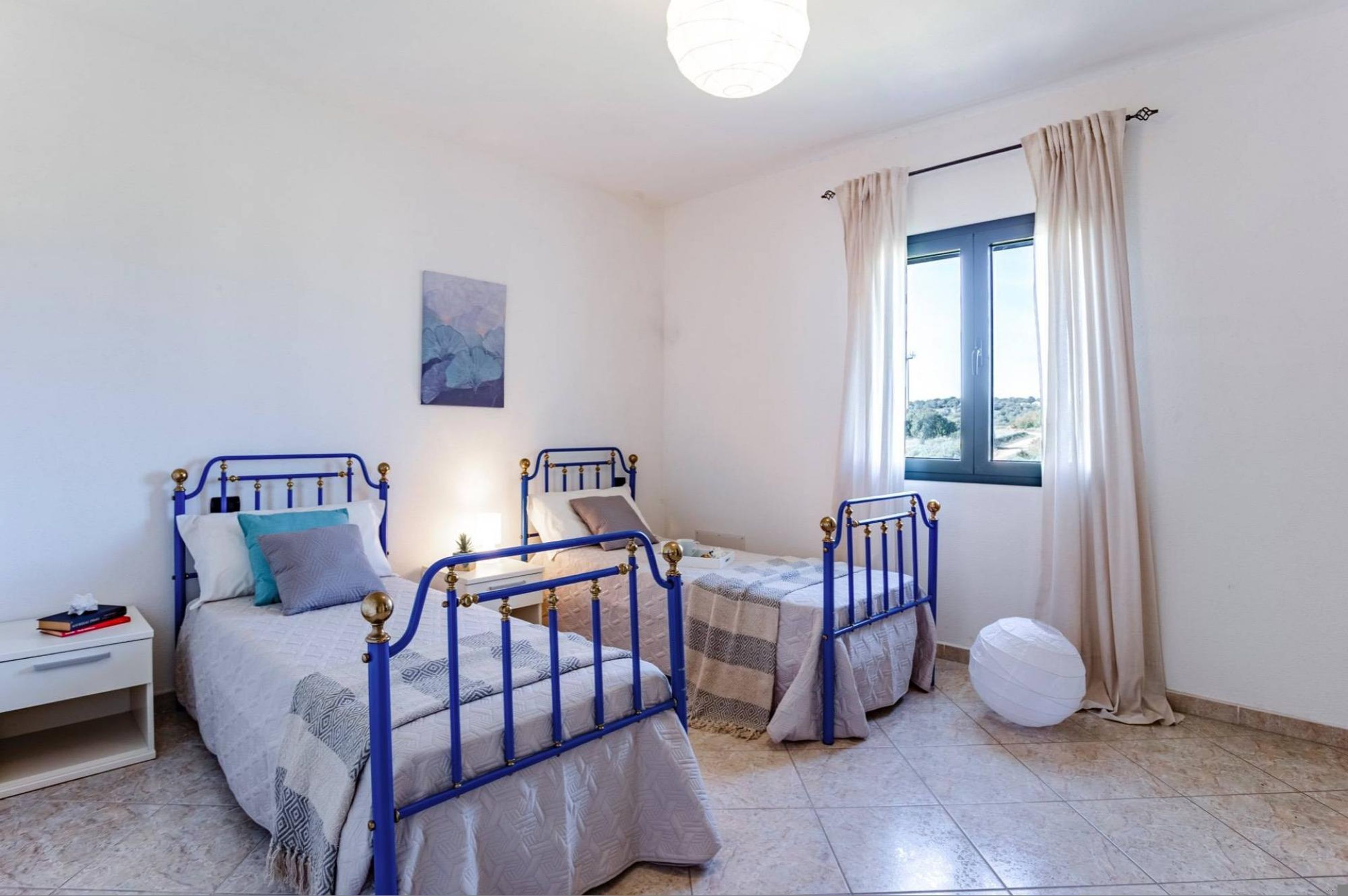 Quarto com duas camas, uma mesa de cabeceira, uma janela com cortinas e um quadro na parede.