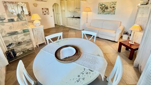 Uma sala de estar acolhedora com uma mesa redonda, cadeiras brancas, um sofá e uma cozinha ao fundo.