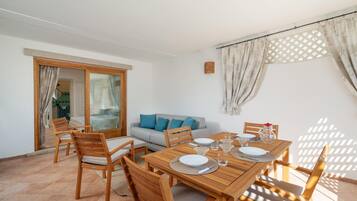 Uma sala de jantar com uma mesa de madeira posta para quatro pessoas, rodeada por cadeiras de madeira. Há um sofá com almofadas azuis ao fundo, e uma porta de vidro deslizante que dá para uma área externa.