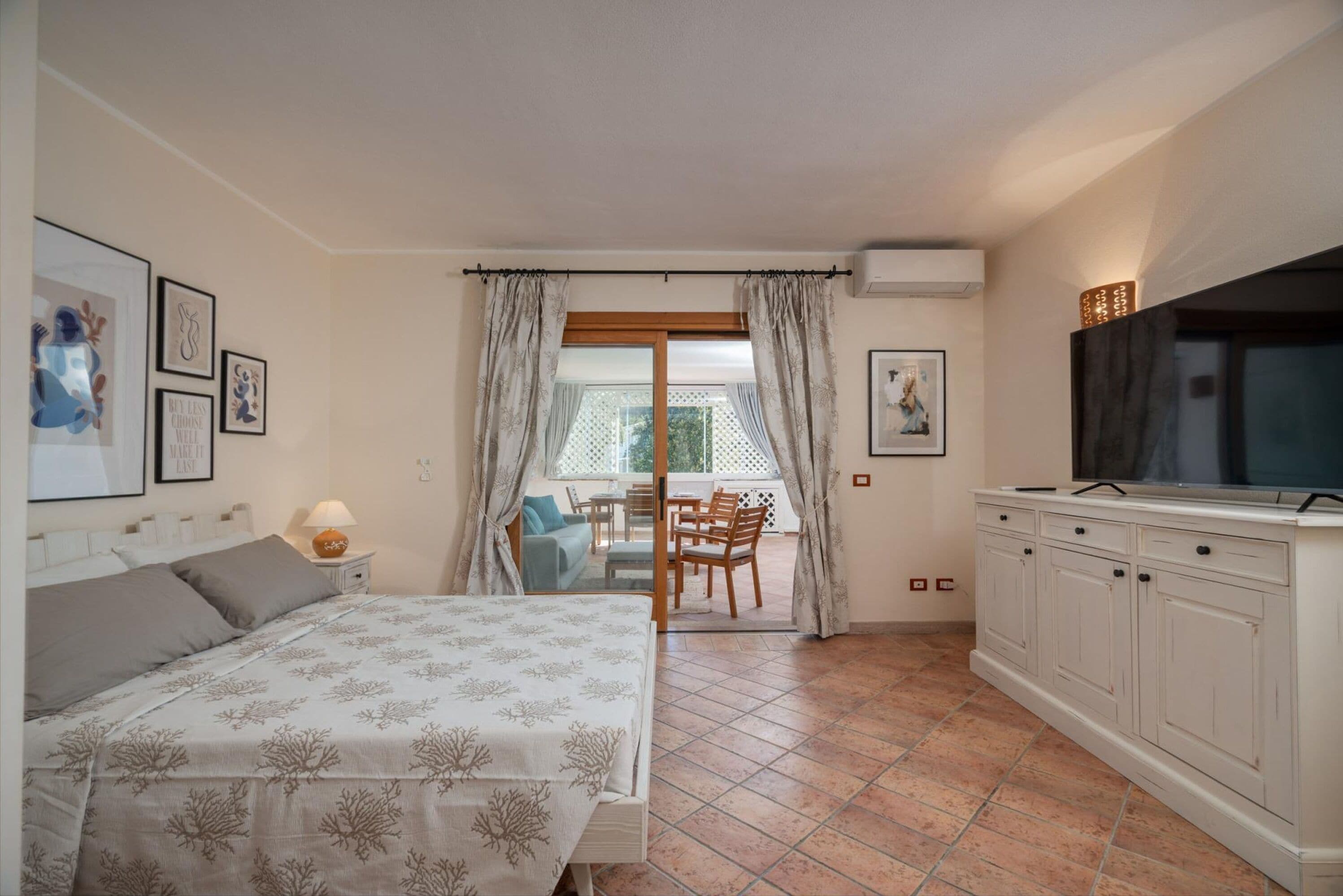 Quarto com cama, televisão, área de refeições com cadeiras e mesa, e varanda com cortinas.