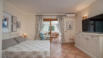 Quarto com cama, televisão, área de refeições com cadeiras e mesa, e varanda com cortinas.
