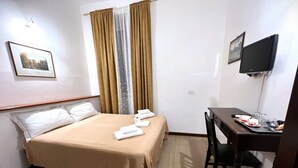 Quarto de hotel com cama, escrivaninha, cadeira, televisão e janela com cortinas.