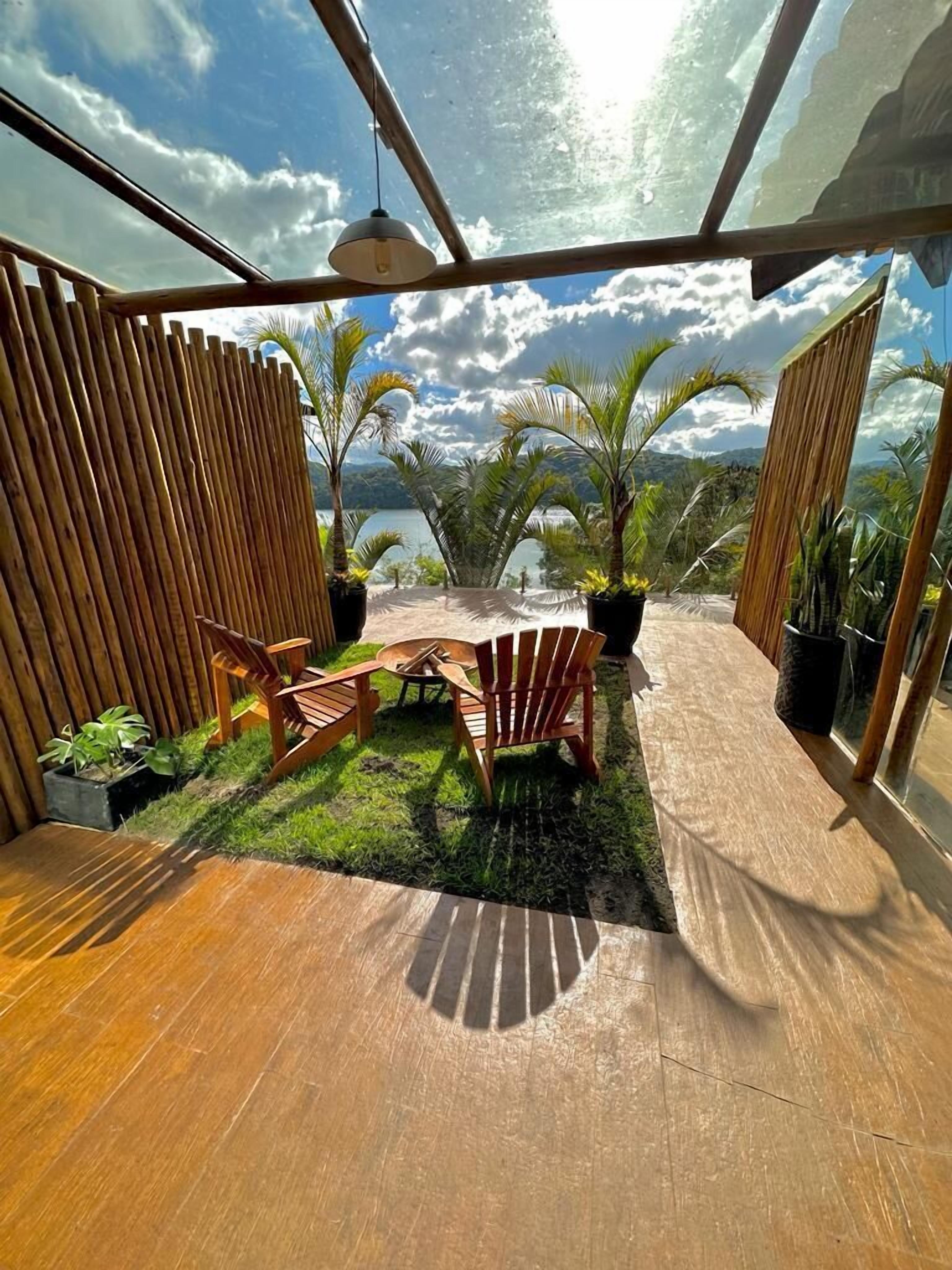 Um pátio moderno ao ar livre com móveis de madeira, teto de vidro e vista para um lago e montanhas.