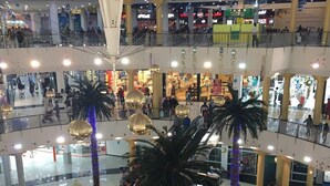 Um shopping center com vários níveis, com escadas rolantes, palmeiras decoradas com luzes azuis e diversas lojas em diferentes andares.
