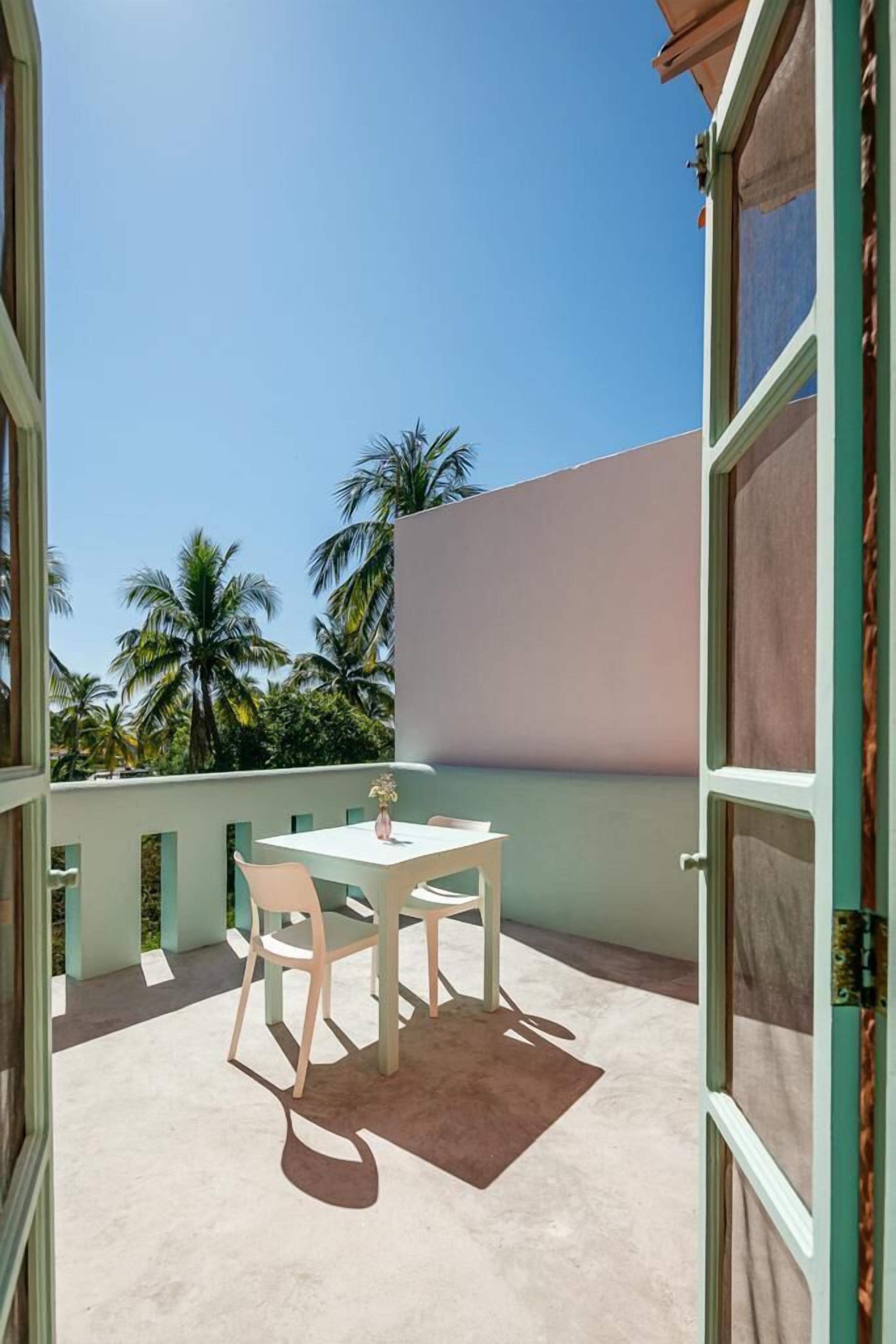 Uma varanda externa com mesa e cadeiras, palmeiras ao fundo e céu claro.