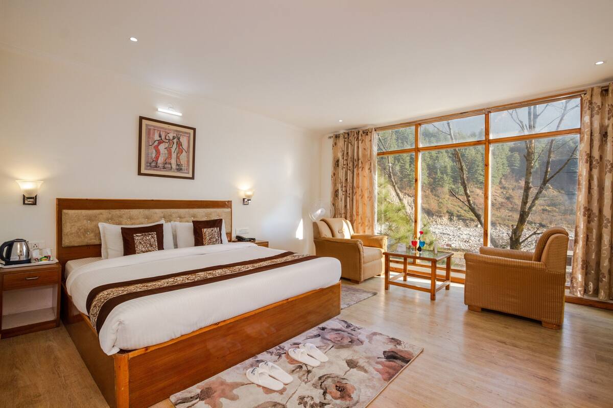 Quarto com cama grande, mesa de cabeceira, cadeira, mesa e janela com vista para árvores.