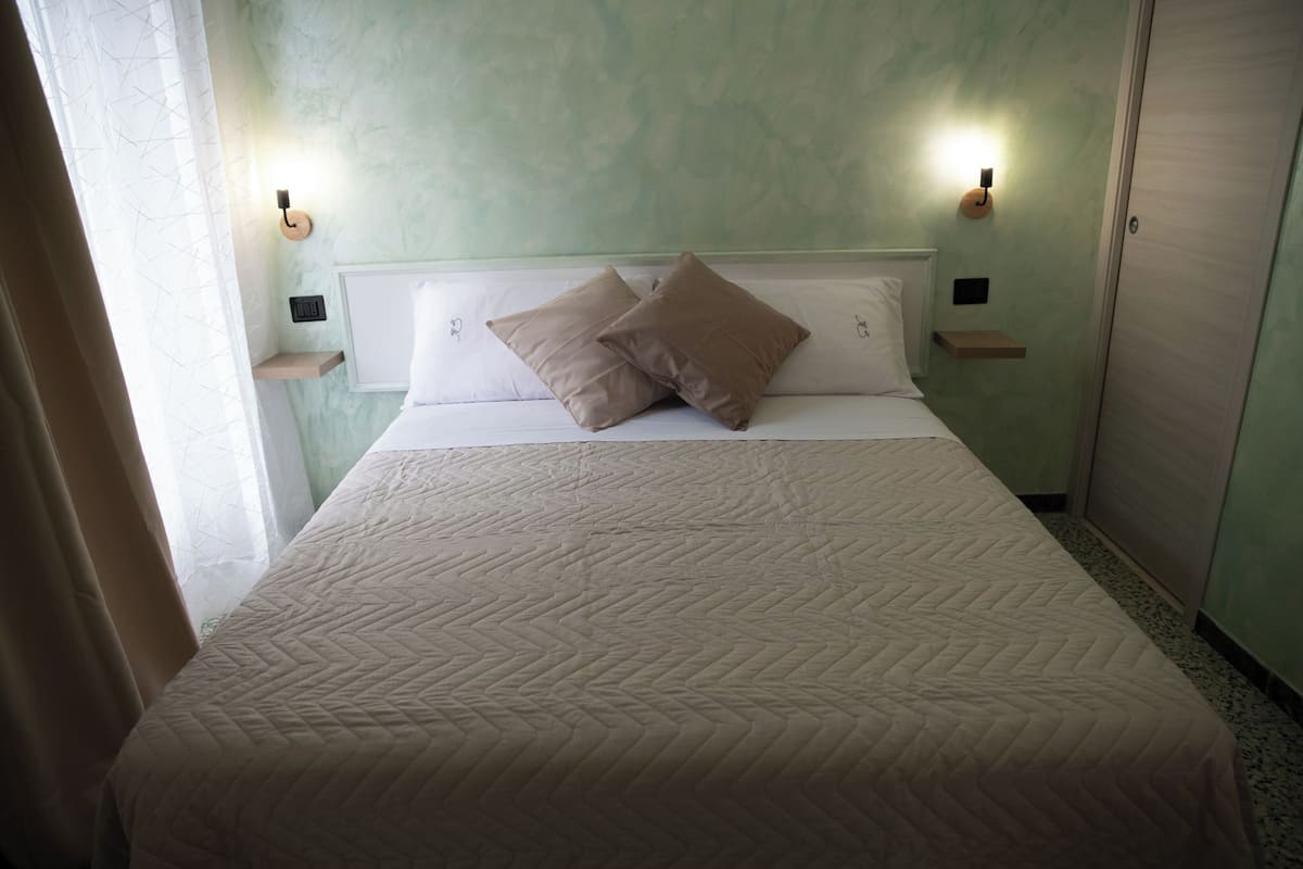 Uma cama bem arrumada com cobertura acolchoada e duas almofadas, uma mesa de cabeceira com abajur, e um candeeiro fixo na parede.
