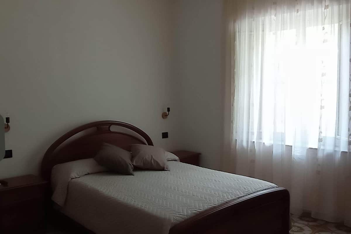 Uma cama de solteiro com cabeceira de madeira, uma mesa de cabeceira e um criado-mudo. Há uma janela com cortinas leves que deixam a luz natural entrar no quarto.