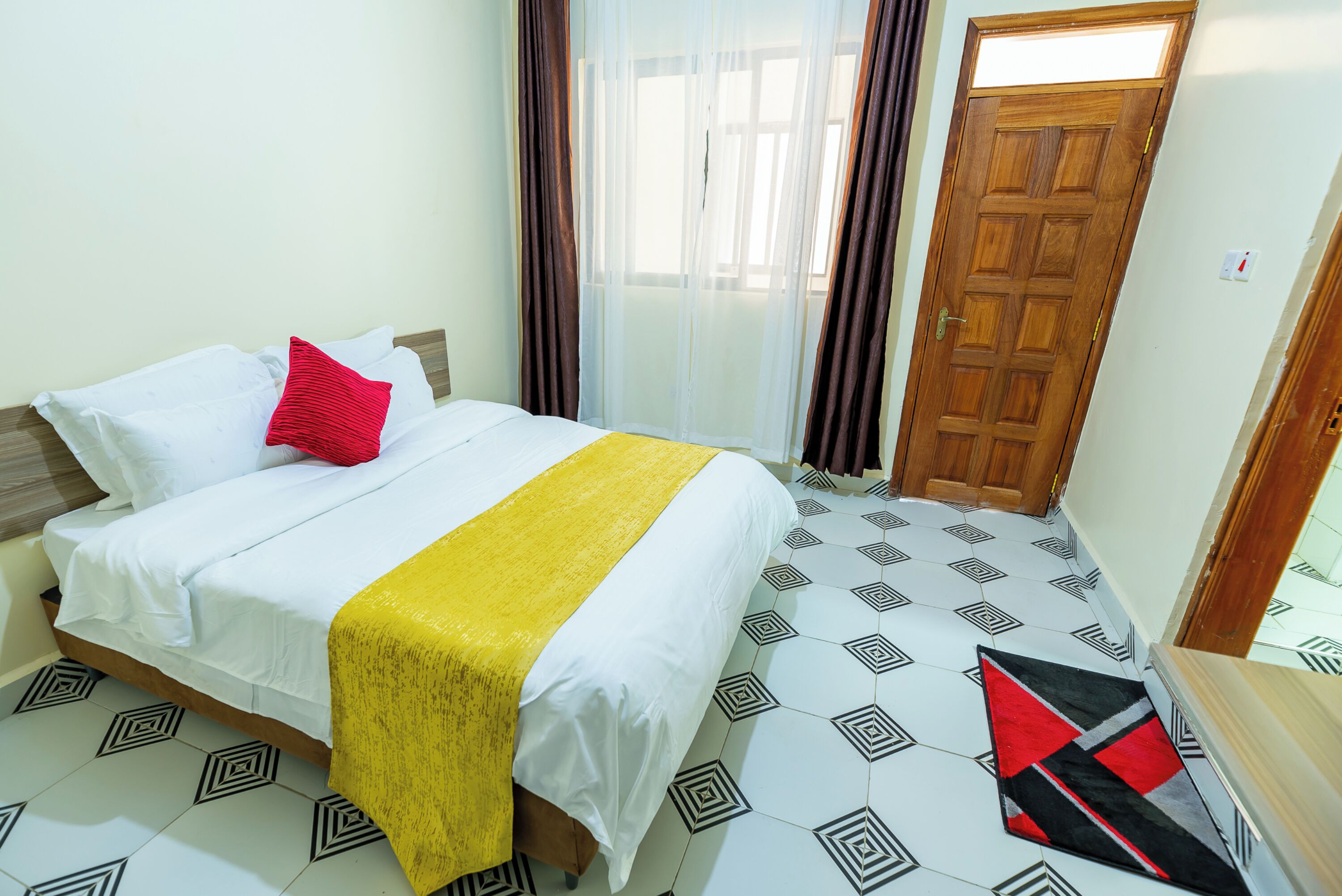 Cama bem arrumada com roupa de cama branca e um cobertor amarelo, um travesseiro vermelho e um chão com padrão geométrico.
