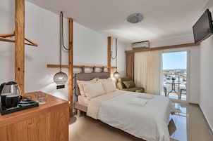 Quarto de hotel com cama, mesa de cabeceira com chaleira e copos, televisão e varanda com vista.