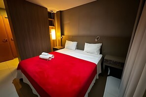 Quarto de hotel com cama, mesa de cabeceira, luminária e guarda-roupa.