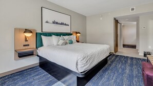 Quarto de hotel com uma cama, duas mesas de cabeceira, uma luminária fixada na parede, uma pintura de paisagem urbana emoldurada e um tapete azul estampado.