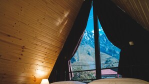 Quarto com teto de madeira, cama com roupa de cama branca e janela com vista para uma paisagem montanhosa.