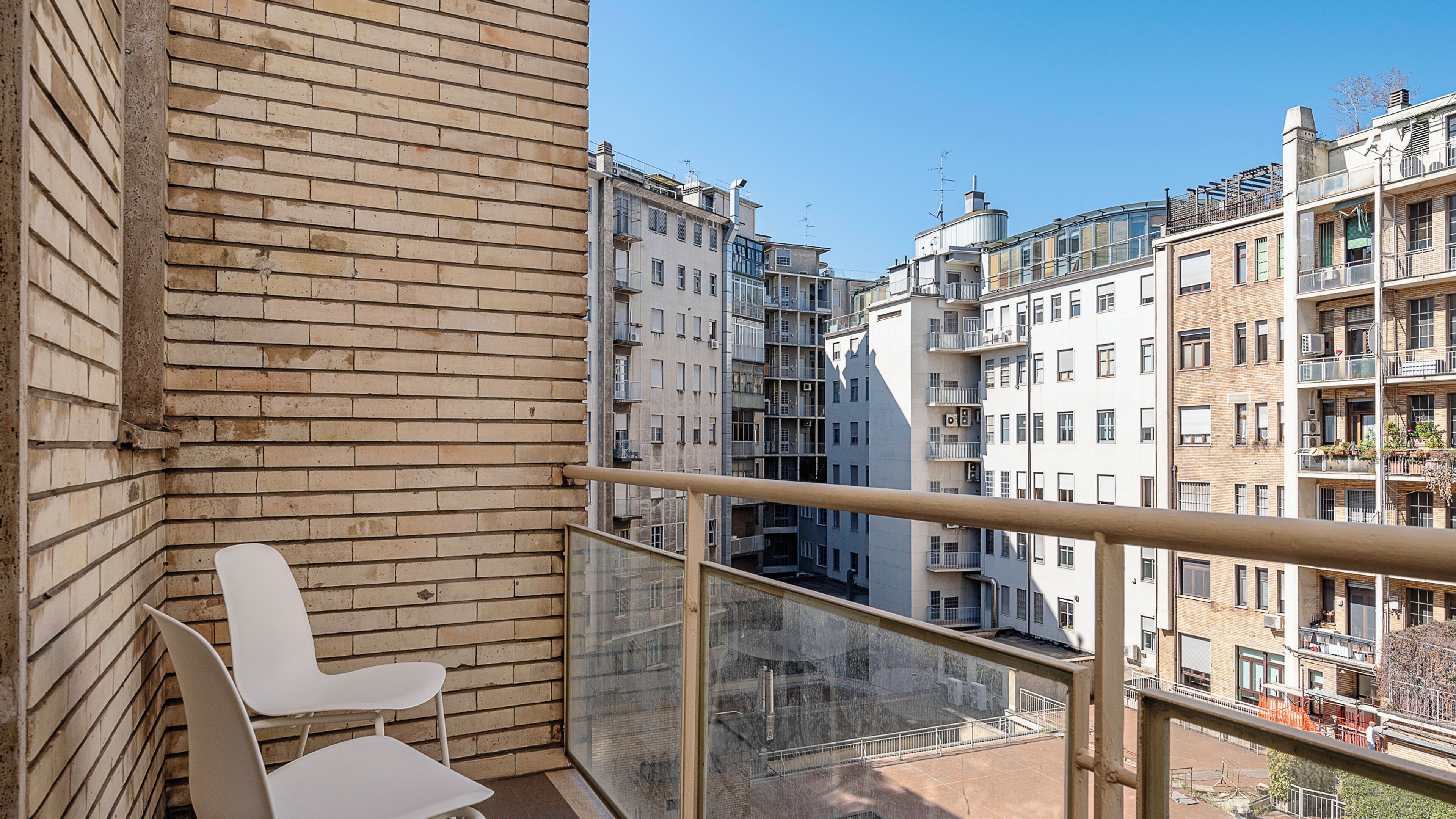 Uma varanda com vista para edifícios próximos, um muro de tijolos e uma cadeira branca.