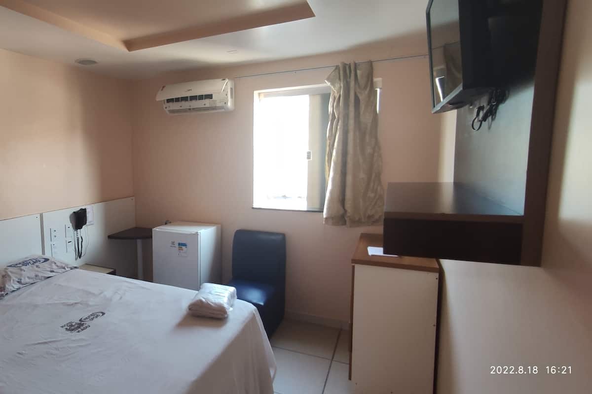 Quarto de hotel com cama, pequena escrivaninha, cadeira, ar condicionado, televisão e janela com cortinas.