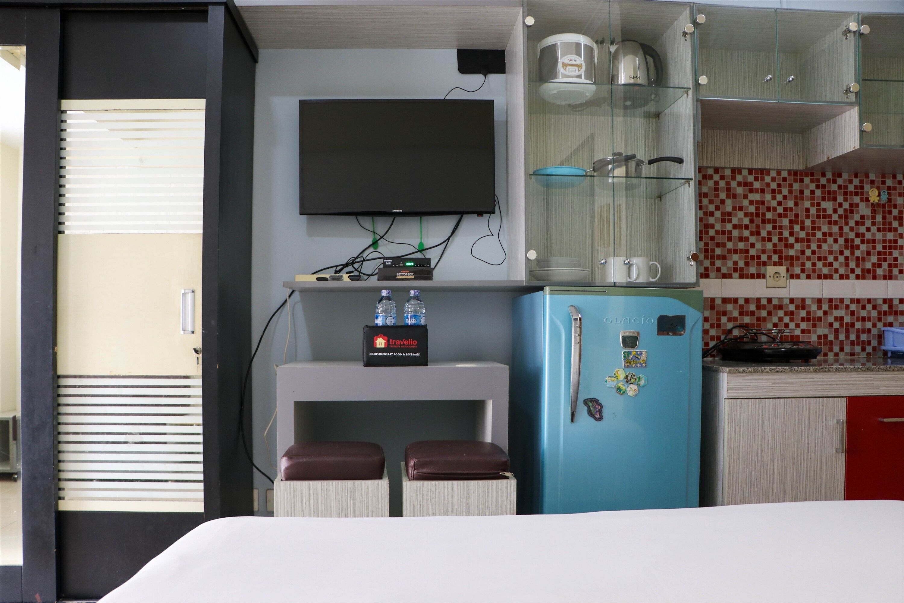 Quarto de hotel compacto com televisão de tela plana sobre uma pequena mesa, geladeira azul e cozinha compacta com painel de azulejos.