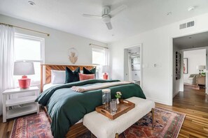 Um quarto com cama, mesas de cabeceira, um criado-mudo, uma janela, um ventilador de teto e uma porta que dá para outro cômodo.