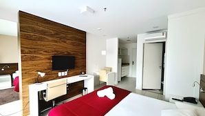 Quarto de hotel moderno com parede de destaque em madeira, televisão de tela plana, escrivaninha com cadeira, cama com colcha vermelha e branca e um guarda-roupa.