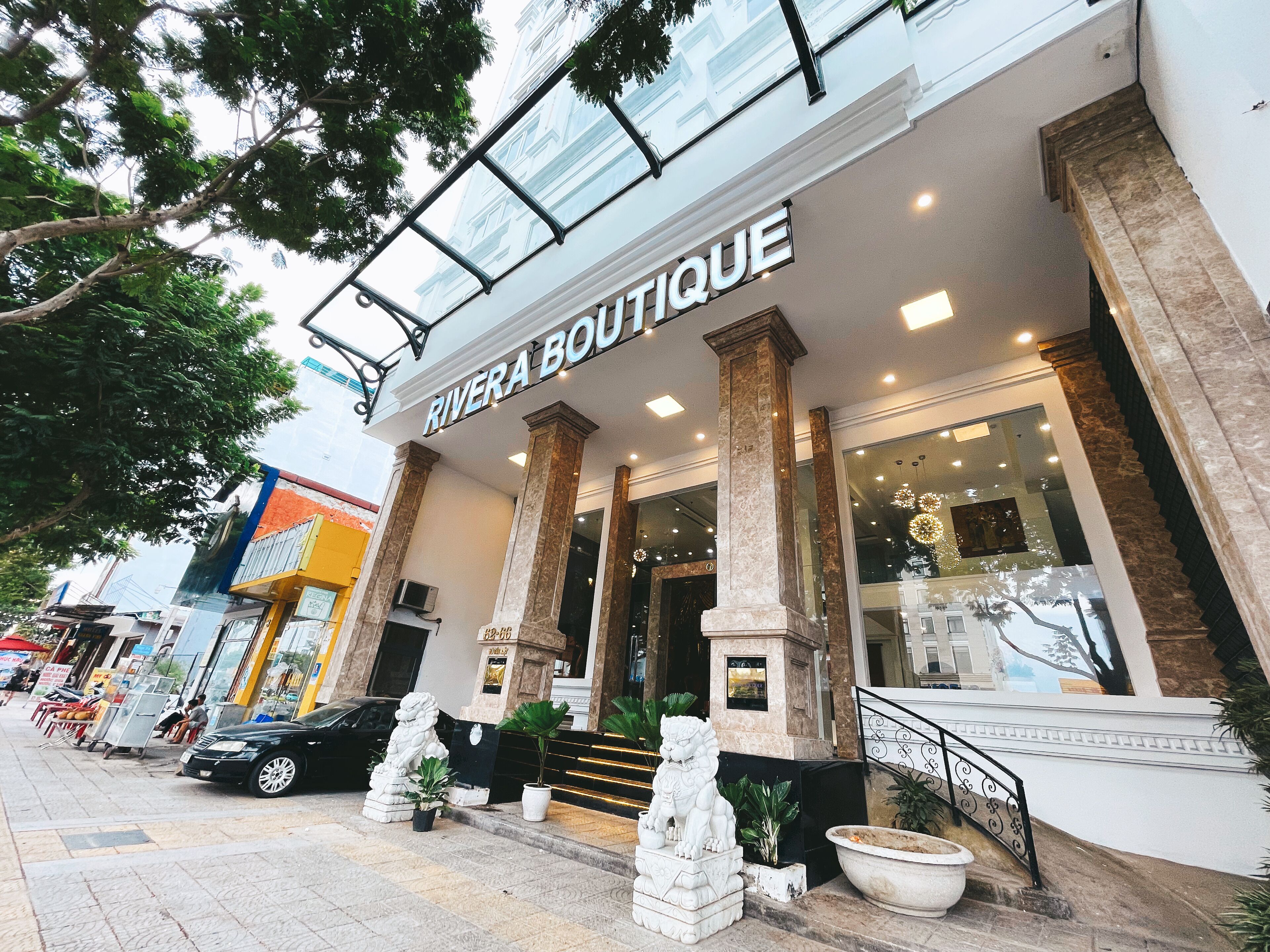 Entrada de um hotel boutique com uma placa indicando "RIVERA BOUTIQUE", apresentando uma fachada imponente com colunas, escadaria e estátua de leão.