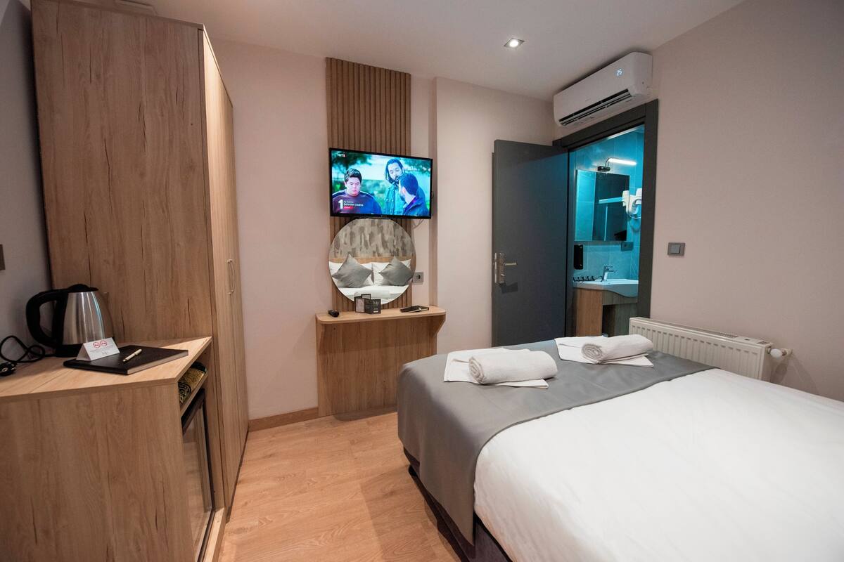 Quarto de hotel com cama, televisão presa à parede, guarda-roupa de madeira, mesa pequena com chaleira e um aviso, e banheiro visível por uma porta aberta.