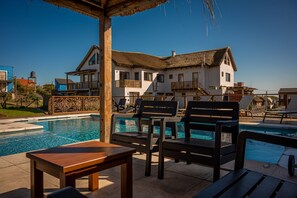 Área da piscina com espreguiçadeiras e uma mesa de madeira, com uma casa de telhado de palha ao fundo, e um céu azul claro.