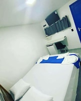 Quarto individual com uma colcha azul, almofadas brancas, uma pequena escrivaninha com cadeira, televisão fixada na parede e porta azul.