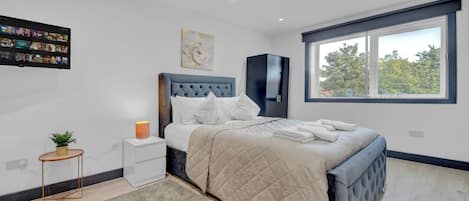 Quarto moderno com uma cama grande, televisão de tela plana, uma mesinha de cabeceira e janela com vista para árvores.