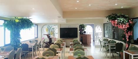 Uma sala de jantar ampla, com várias mesas e cadeiras, uma televisão de tela plana e plantas decorativas.