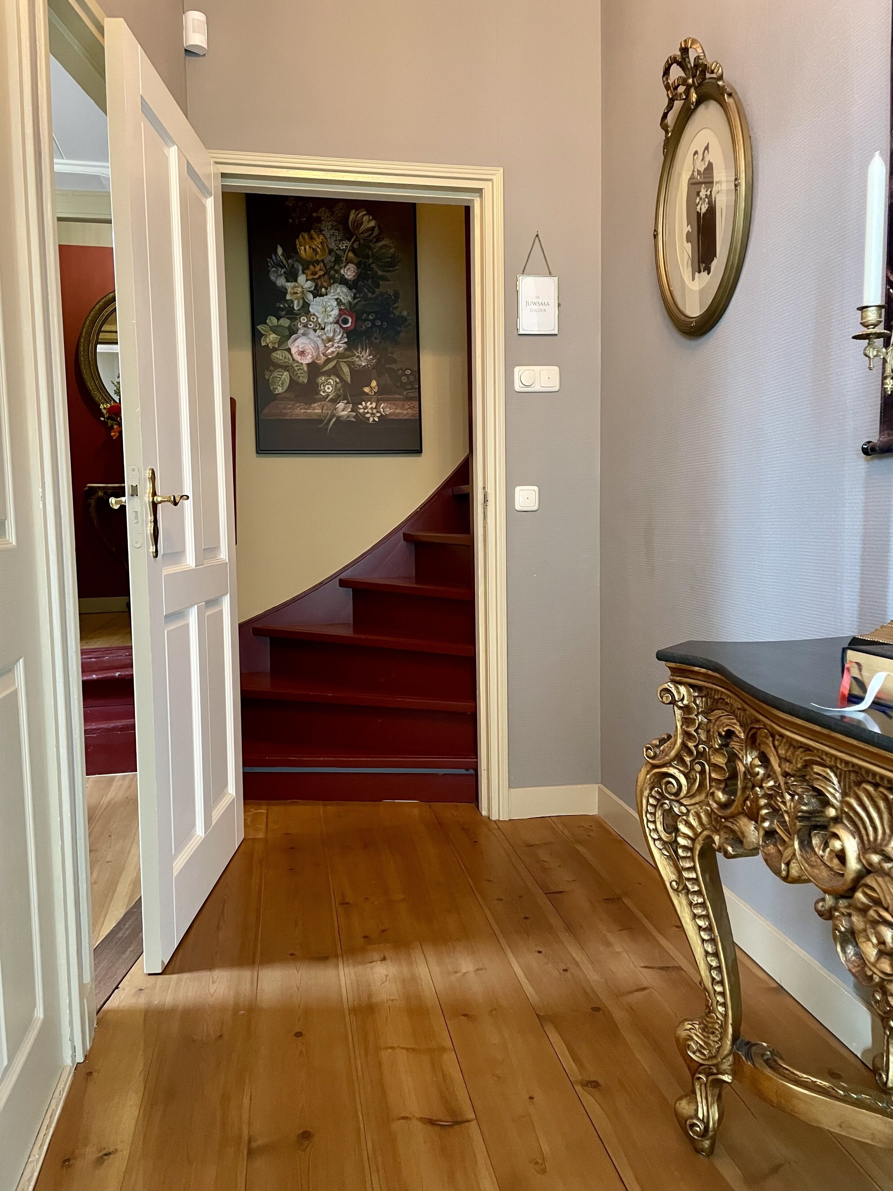 Um corredor com piso de madeira, escada vermelha, um quadro, um espelho e um console decorativo.