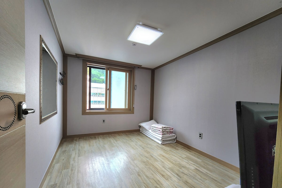 Quarto com uma janela grande, piso de madeira, um espelho e uma televisão.