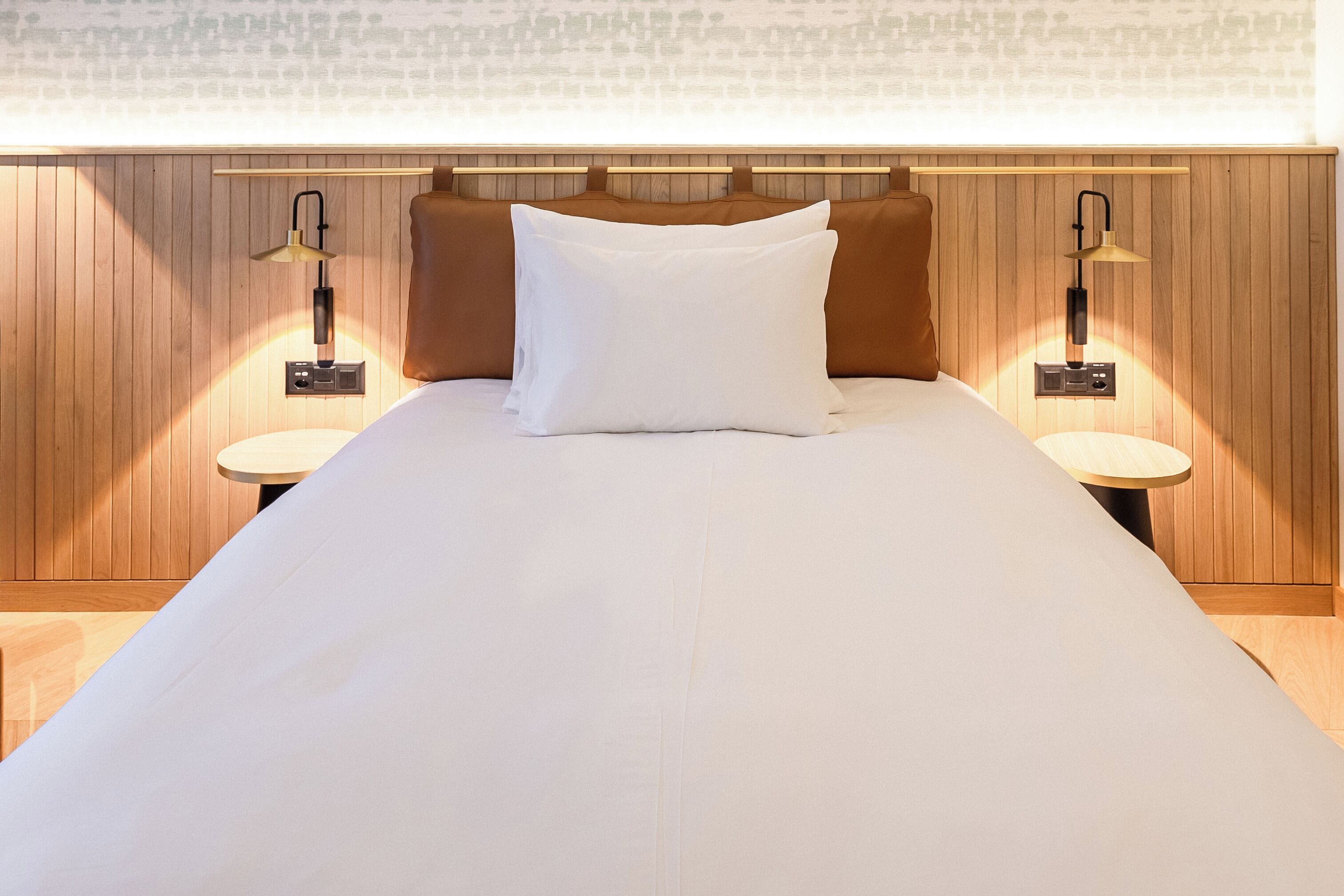 Cama bem arrumada com lençóis brancos e duas almofadas castanhas, com duas mesas de cabeceira com luminárias, num quarto com painéis de madeira e parede com padrão de tijolos.