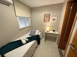 Quarto pequeno e bem arrumado com cama, mesa de cabeceira com abajur, janela com persianas e porta.