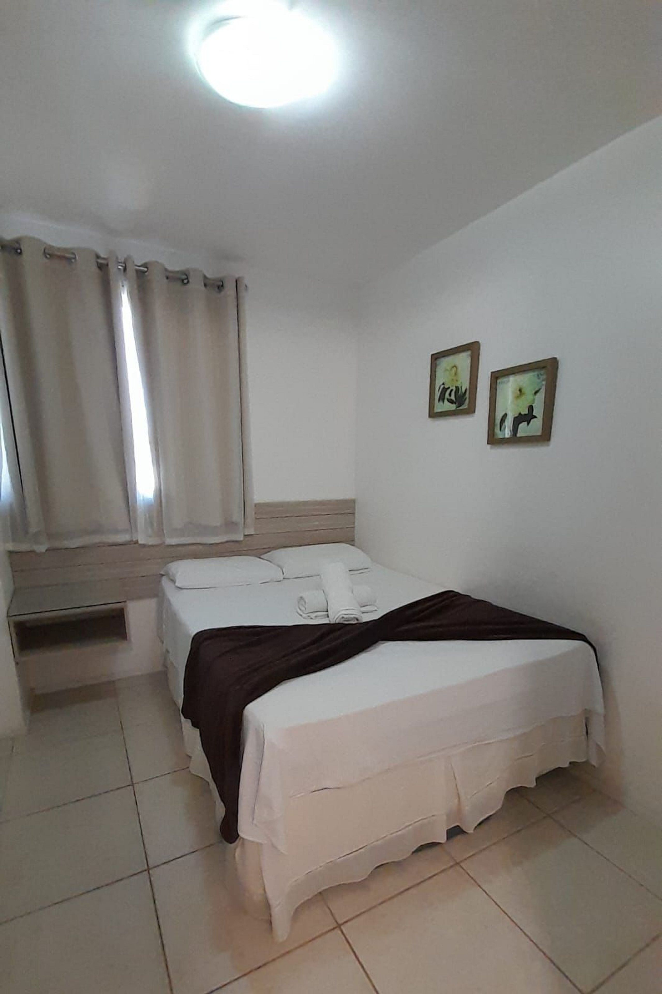 Quarto de hotel com cama, janela com cortinas, cabeceira e duas fotos emolduradas na parede.