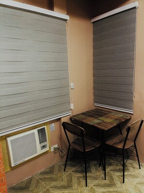 Quarto de canto com uma mesa, cadeiras, um radiador e duas janelas grandes com persianas.