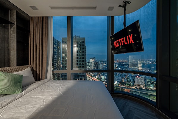 Quarto com cama grande, vista para a cidade através de uma janela ampla e placa indicando que o quarto está disponível para Netflix.