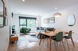 Uma sala de estar moderna com uma mesa de jantar de madeira, cadeiras verdes, um sofá cinza e uma janela grande.