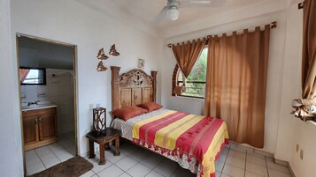 Quarto com cama, cabeceira de madeira, janela com cortinas, mesa de cabeceira e espelho.