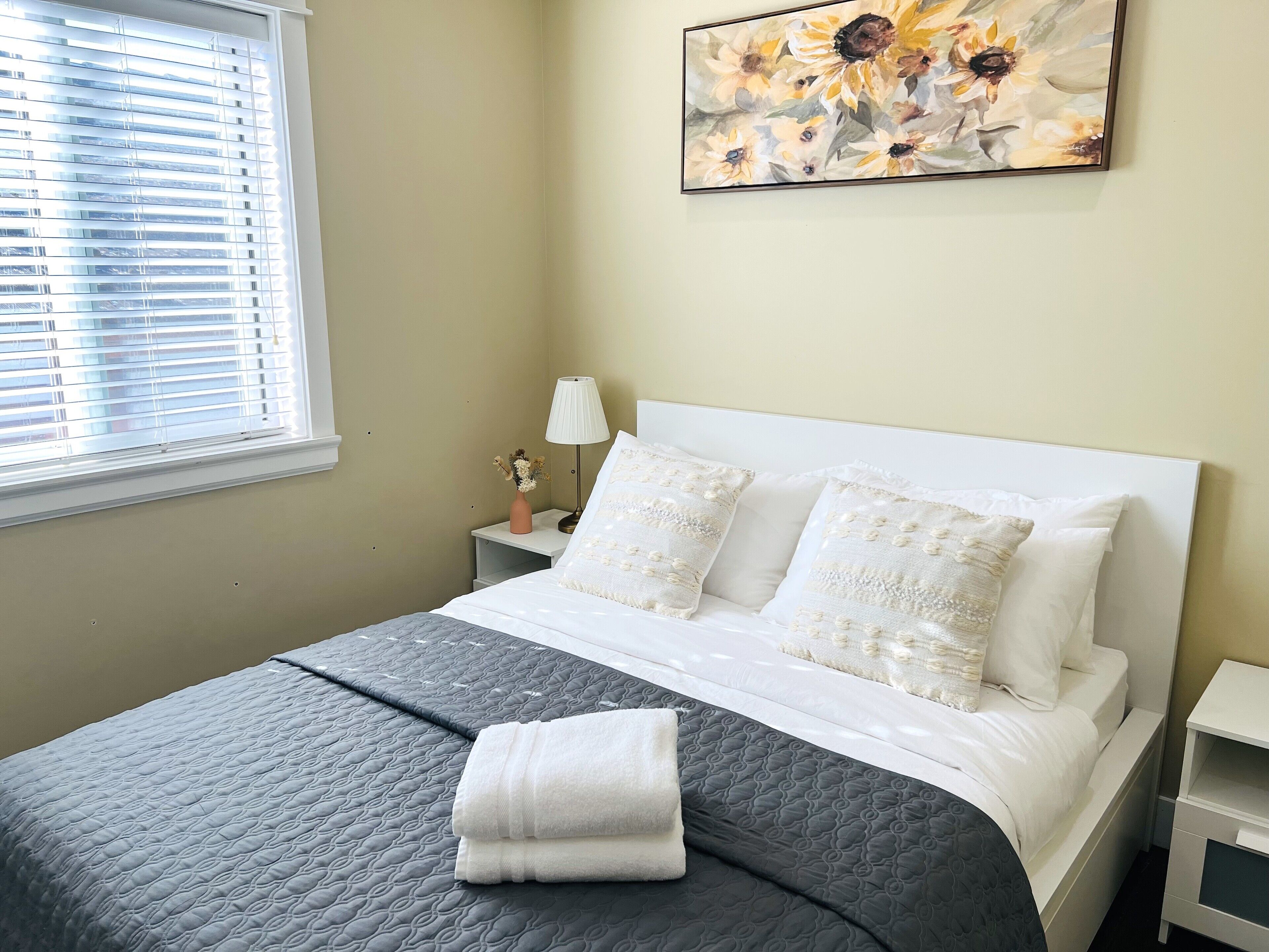 Cama bem arrumada com colcha cinza, almofadas brancas e toalha branca ao lado. Um quadro com flores está pendurado na parede acima da cama.