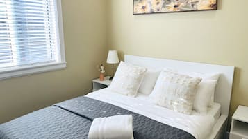 Cama bem arrumada com colcha cinza, almofadas brancas e toalha branca ao lado. Um quadro com flores está pendurado na parede acima da cama.