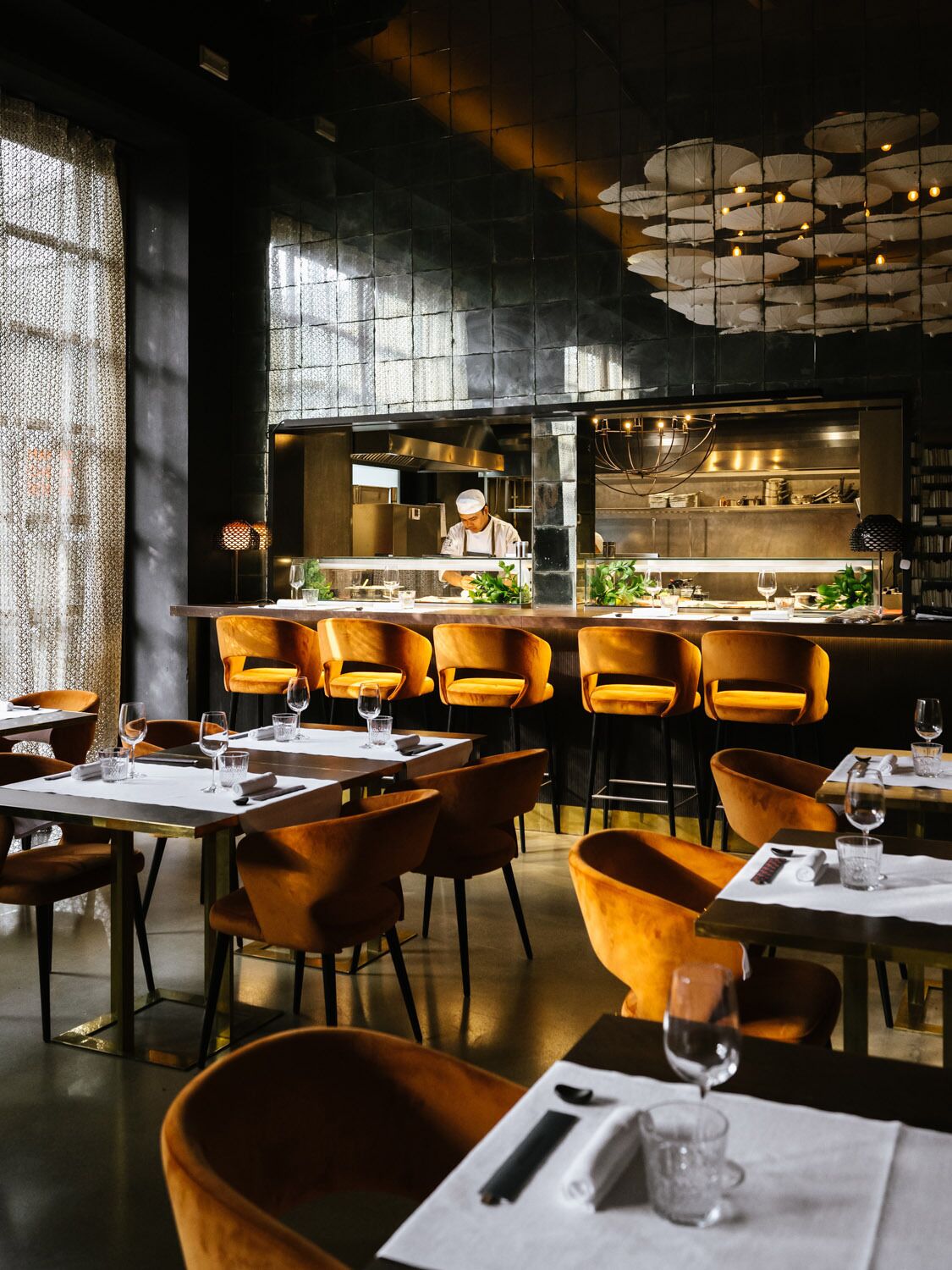Interior de restaurante com um chef preparando comida, mesas e cadeiras de madeira e um lustre moderno.