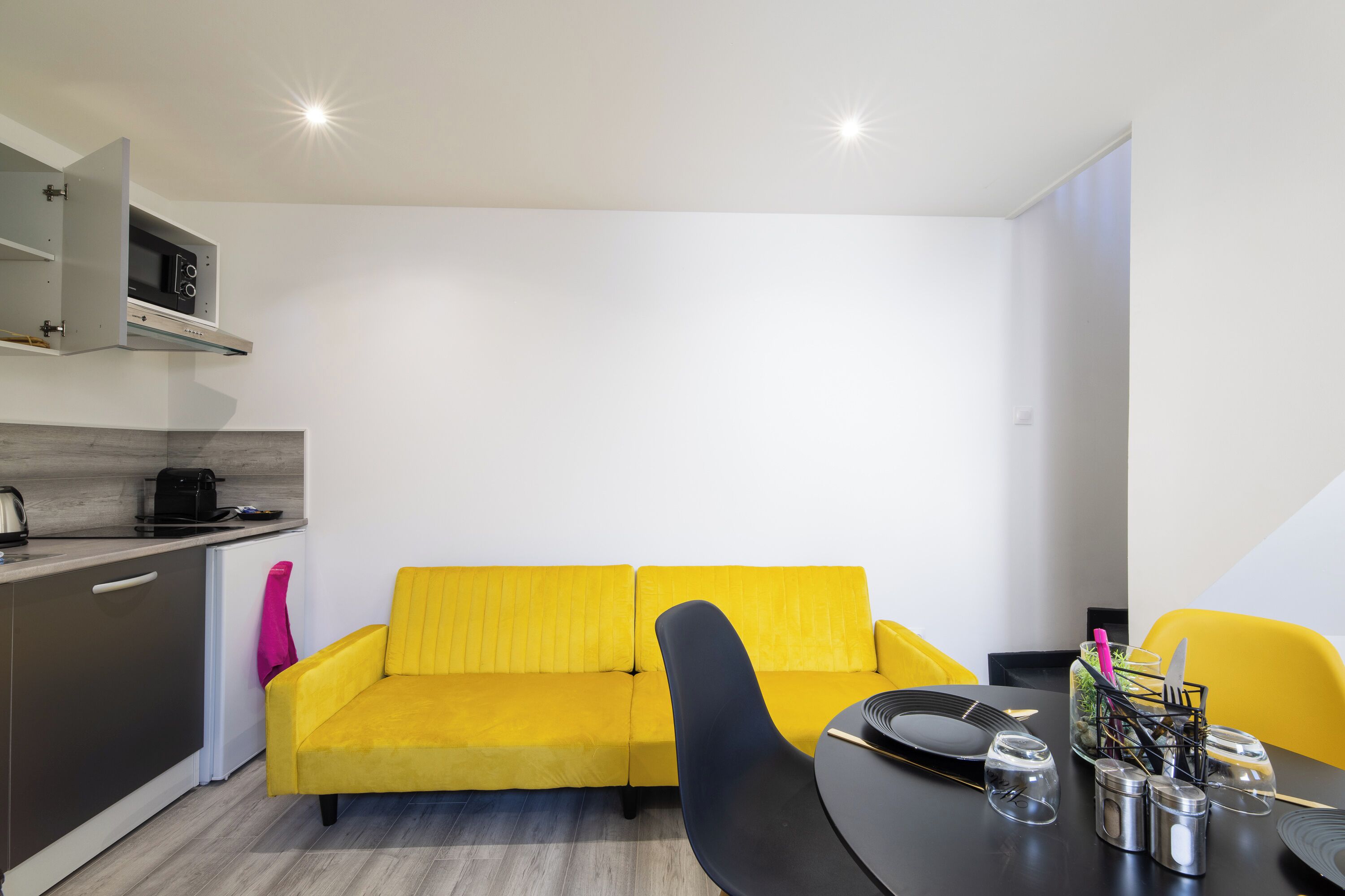 Um espaço de estar compacto com cozinha compacta, um sofá amarelo, uma mesa de jantar e uma cadeira preta.