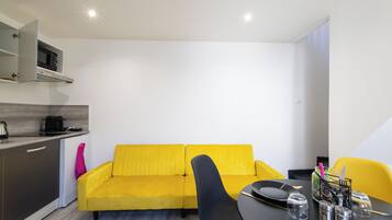 Um espaço de estar compacto com cozinha compacta, um sofá amarelo, uma mesa de jantar e uma cadeira preta.