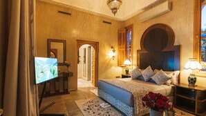 Quarto de hotel com uma cama grande, televisão, mesa de cabeceira com abajur, um vaso com flores e cabeceira de madeira.