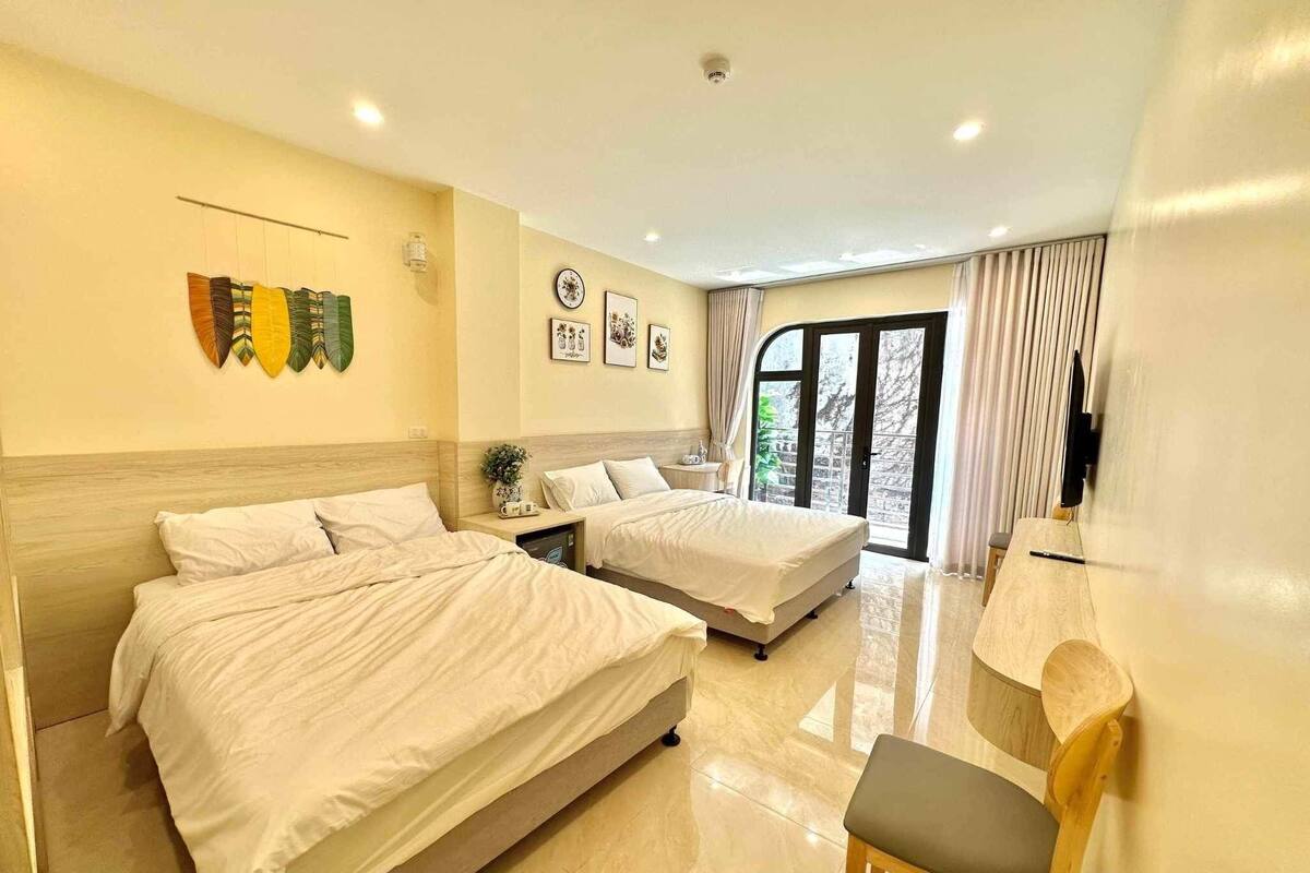 Quarto de hotel com duas camas, uma escrivaninha, uma cadeira, televisão e varanda com cortinas.