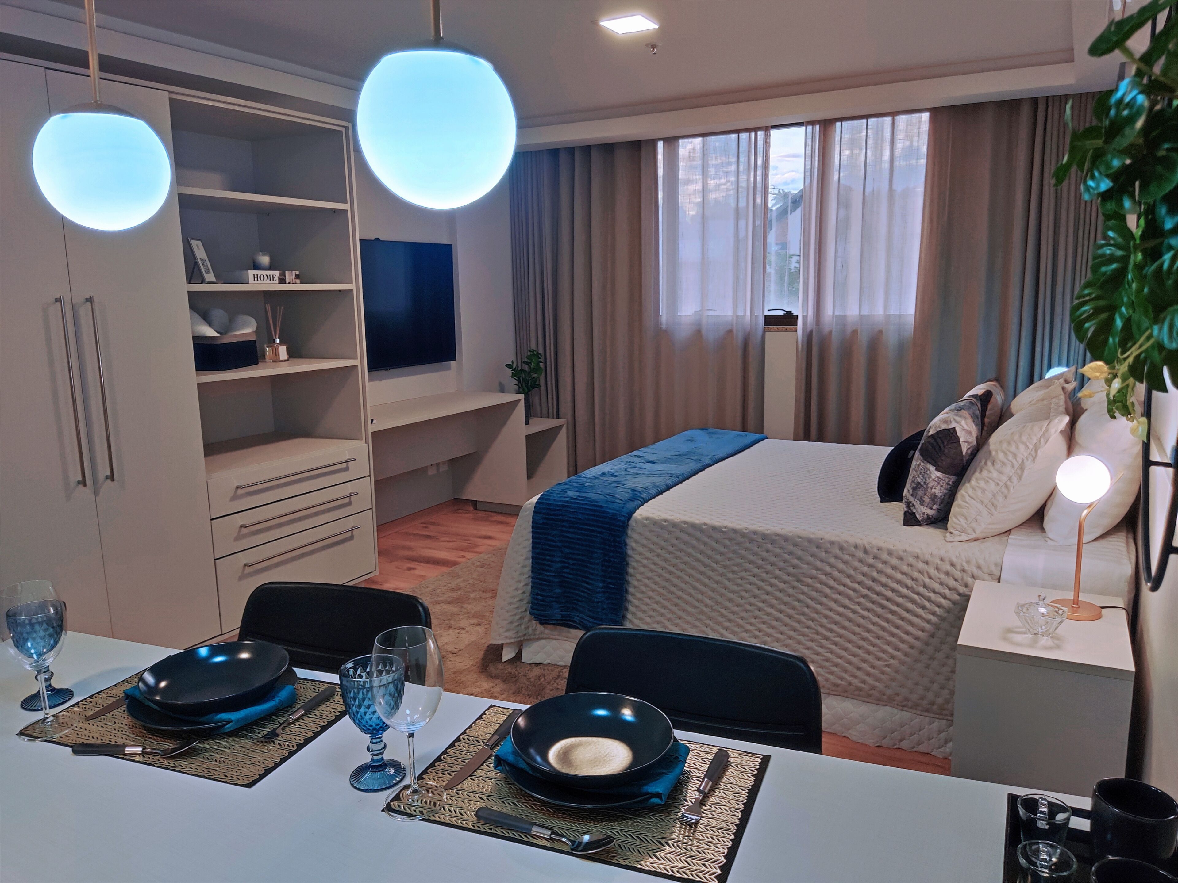 Quarto de hotel moderno com uma cama bem arrumada, área de jantar com mesa posta para dois e televisão na parede.