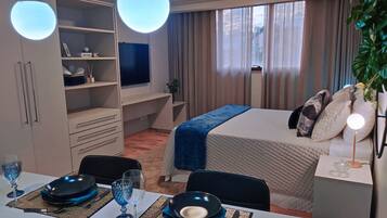 Quarto de hotel moderno com uma cama bem arrumada, área de jantar com mesa posta para dois e televisão na parede.